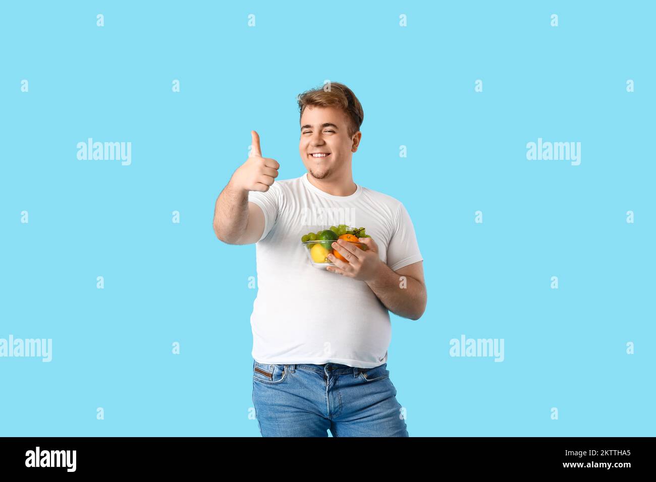 Young overweight man with bowl of fruits showing thumb-up on blue ...