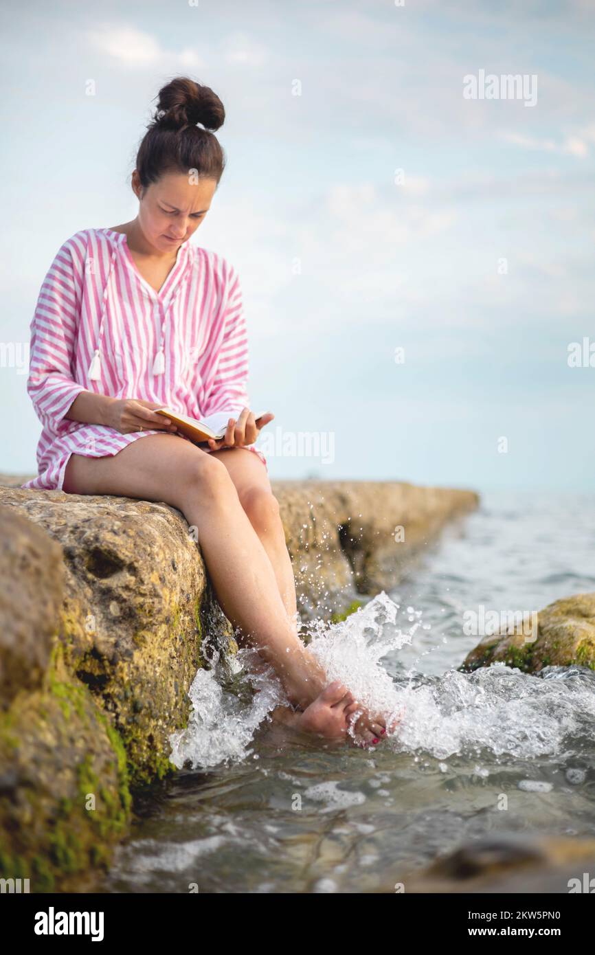 Happy travel woman reading book sitting on dock pier legs in water ...