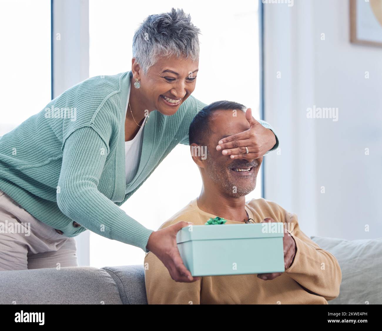 Senior couple, gift and surprise in home living room with wife covering ...