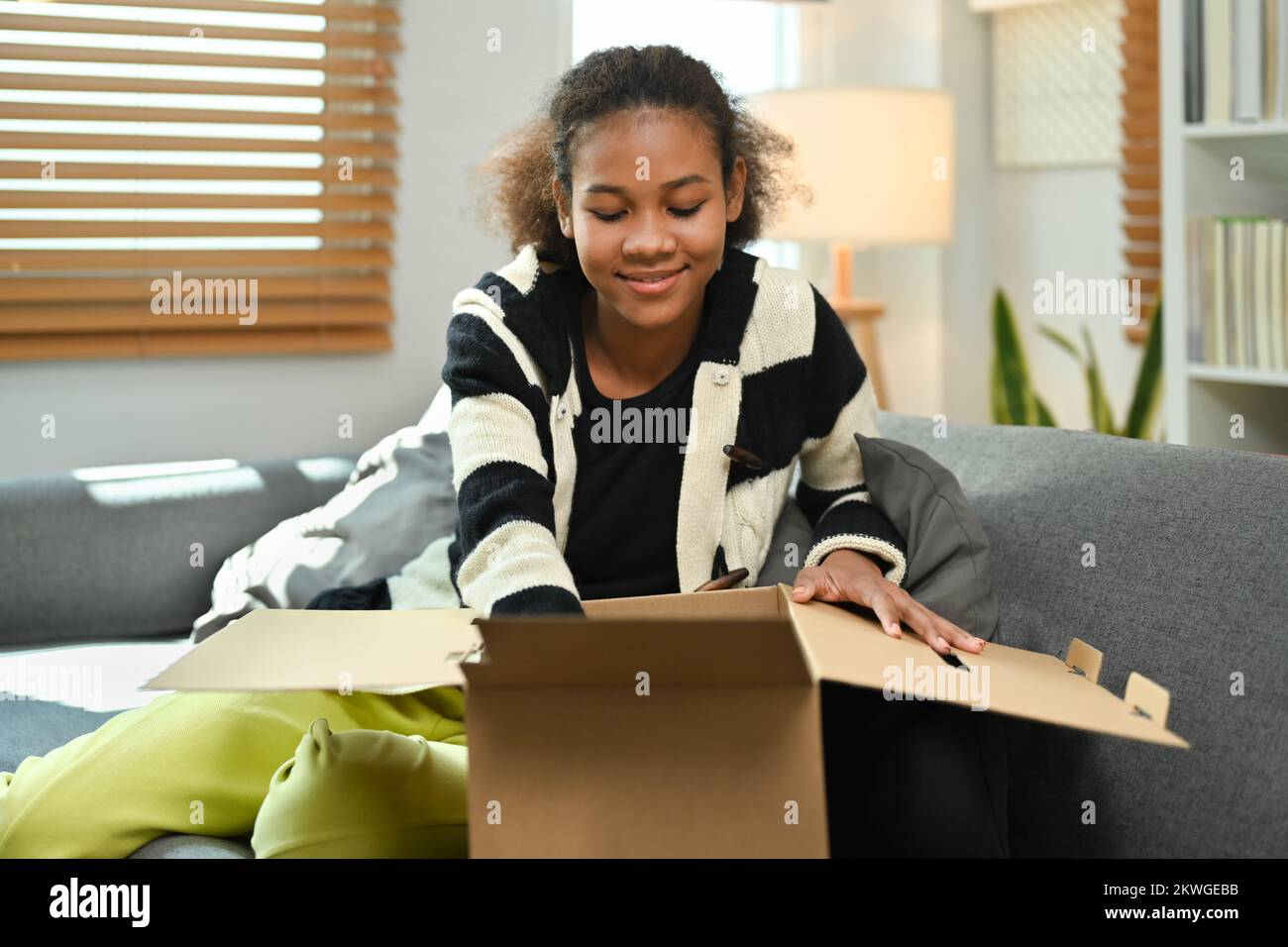 Pleased young African American woman sitting on couch and unpacking ...