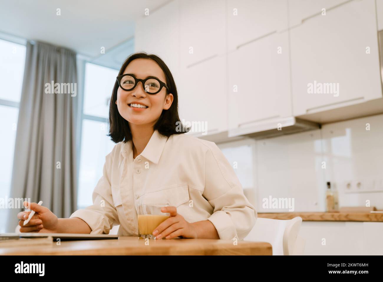 Young cute smiling asian woman in glasses working by table in kitchen ...