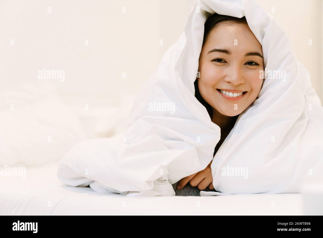Young smiling asian girl in home suit lying on bed under covers at home ...