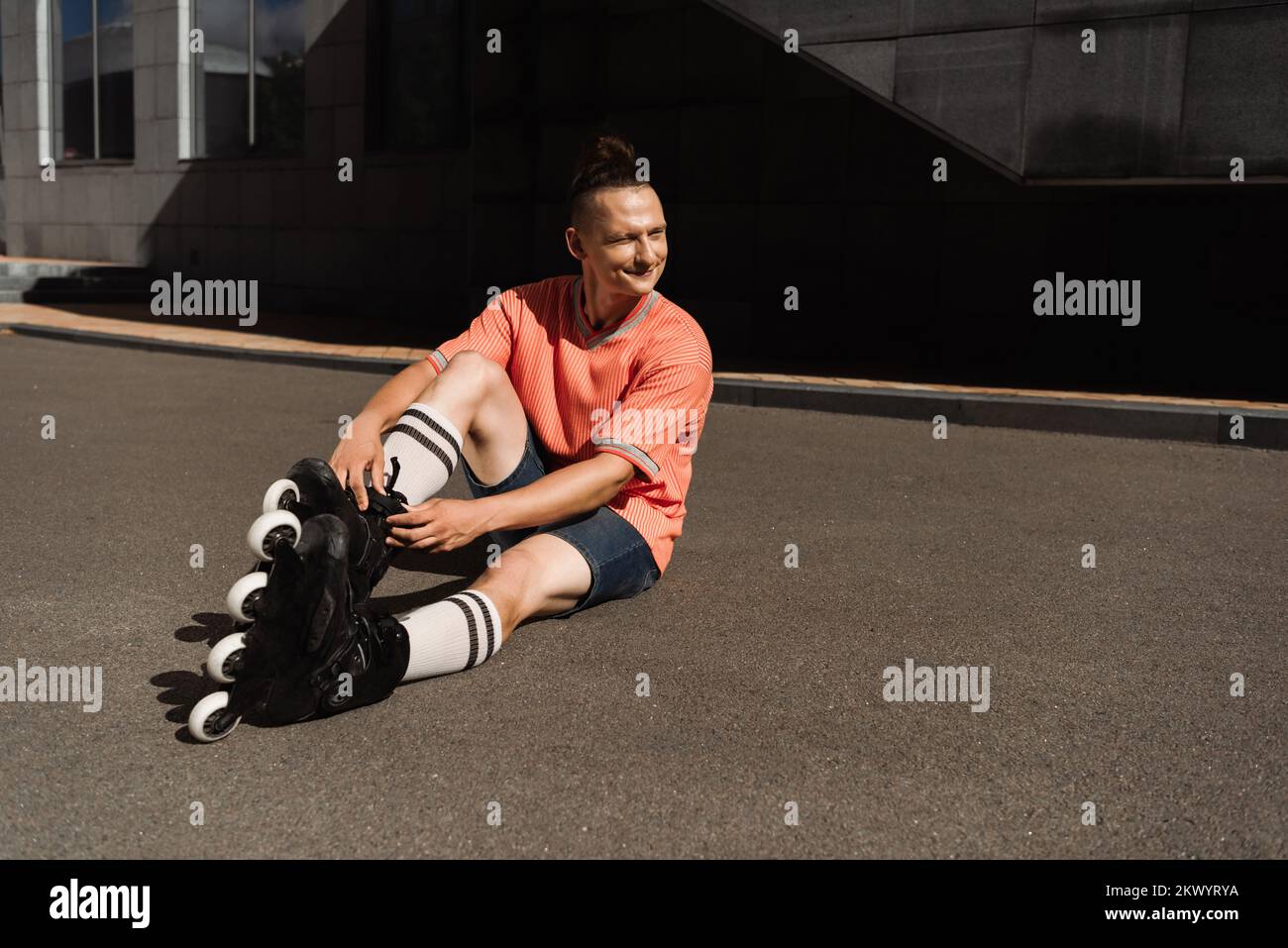 Positive man wearing rollers while sitting on asphalt on urban street ...