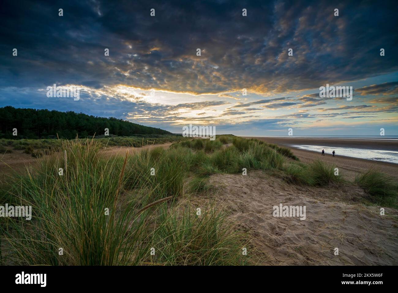A couple walk along the beach and sand dunes during sunset at Holkham ...
