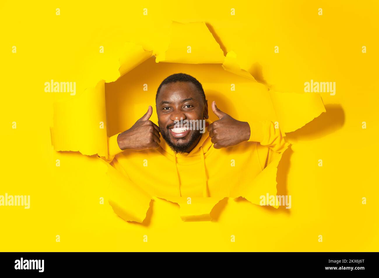 Joyful black man gesturing thumbs up and smiling at camera through hole ...