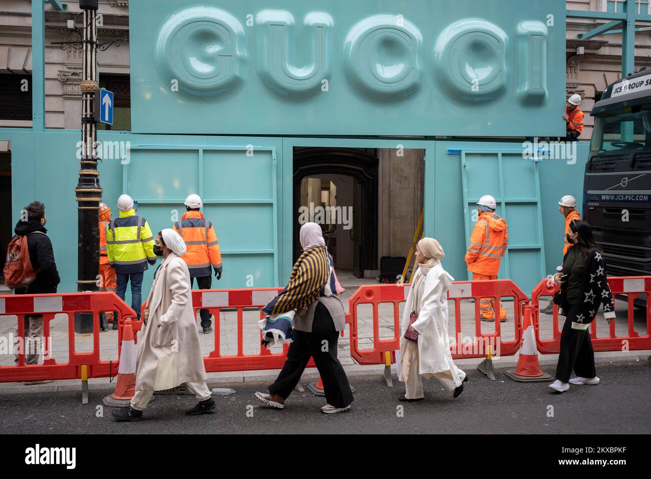 A family walk past workmen preparing to position a Gucci logo sign, to ...