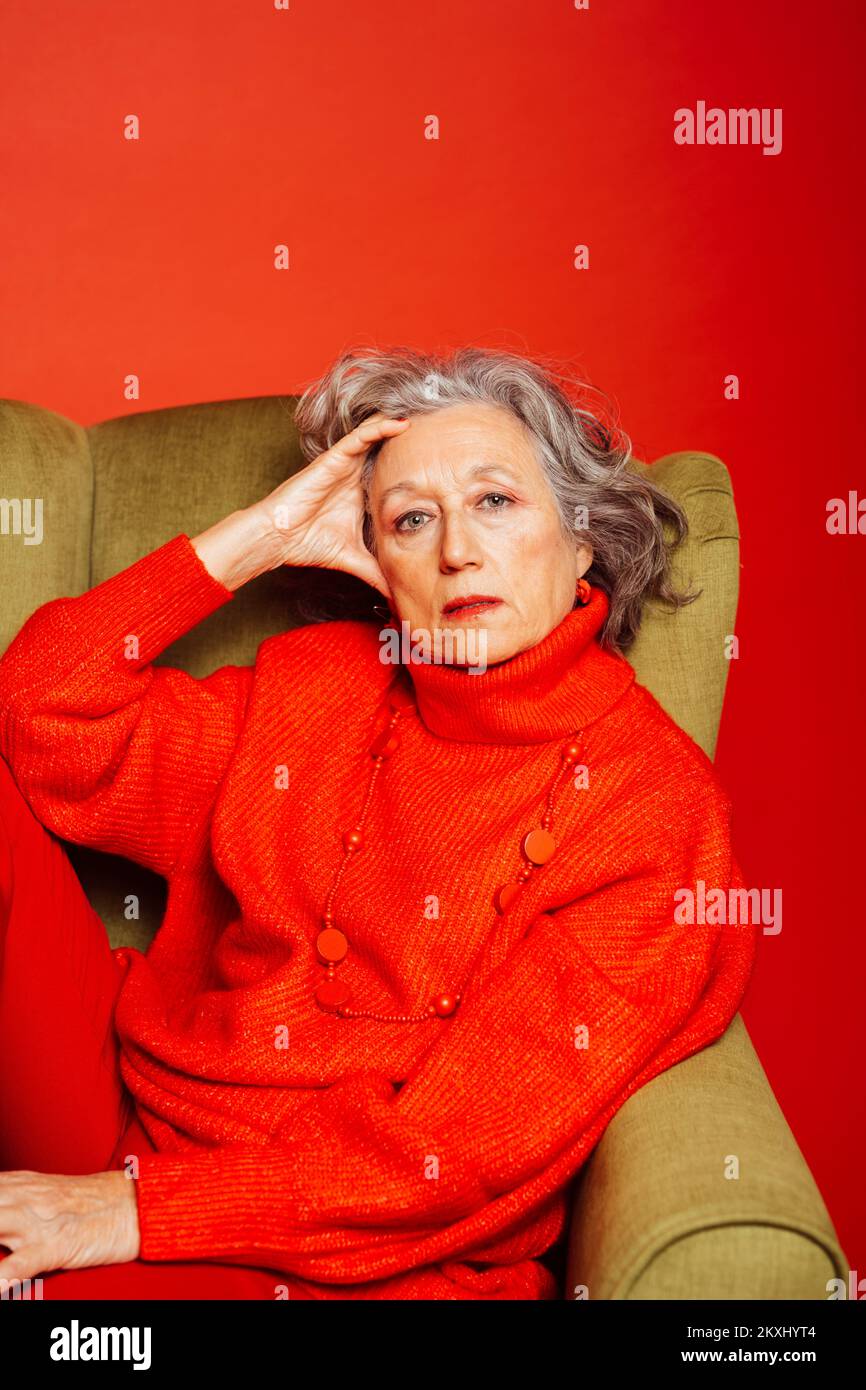 Senior woman wearing red clothes and sitting on a green armchair over a ...