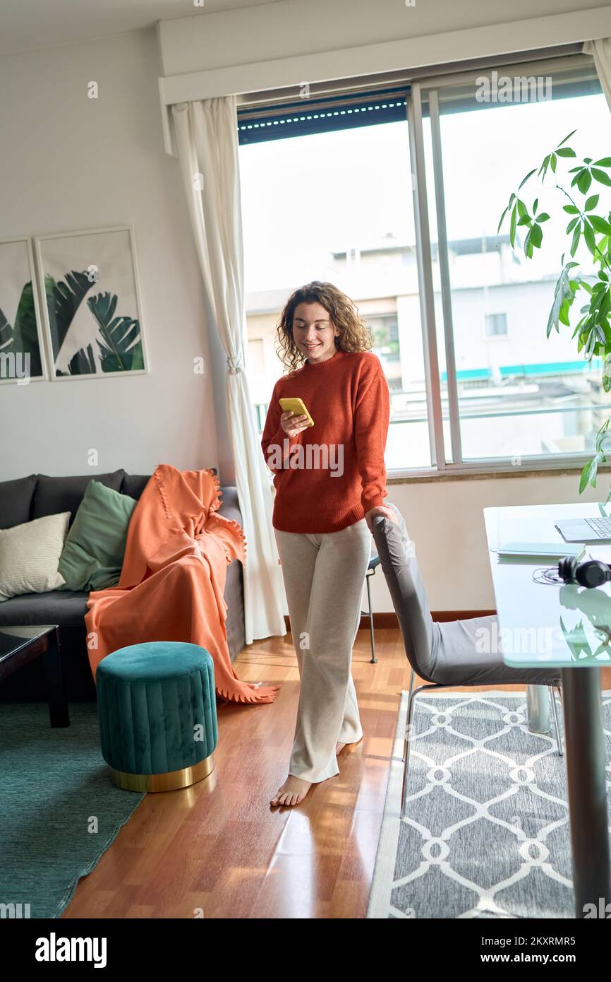 Smiling young woman using cell phone standing in cozy living room at ...