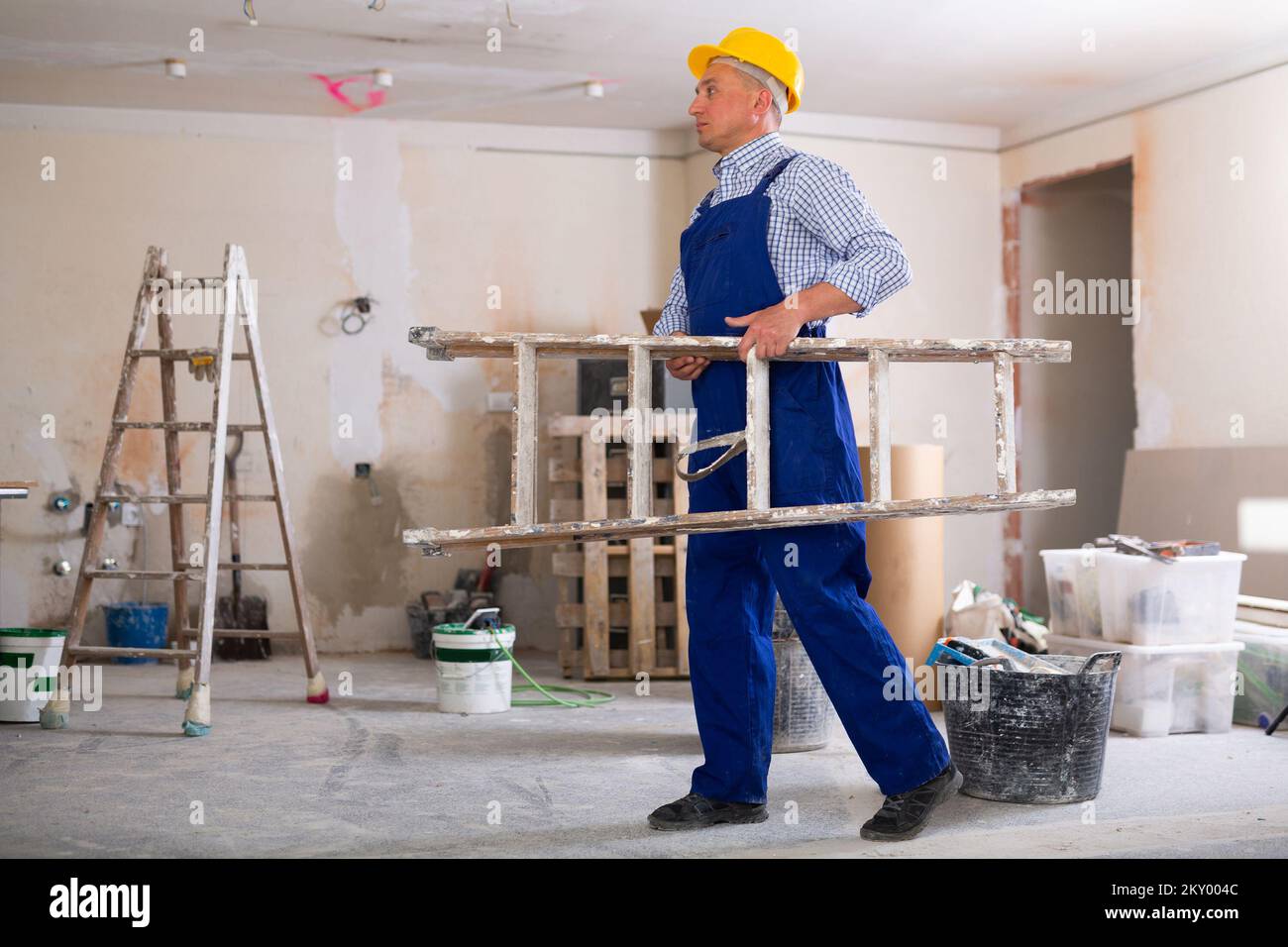 Worker in blue overalls carries a ladder on his shoulder in room being ...