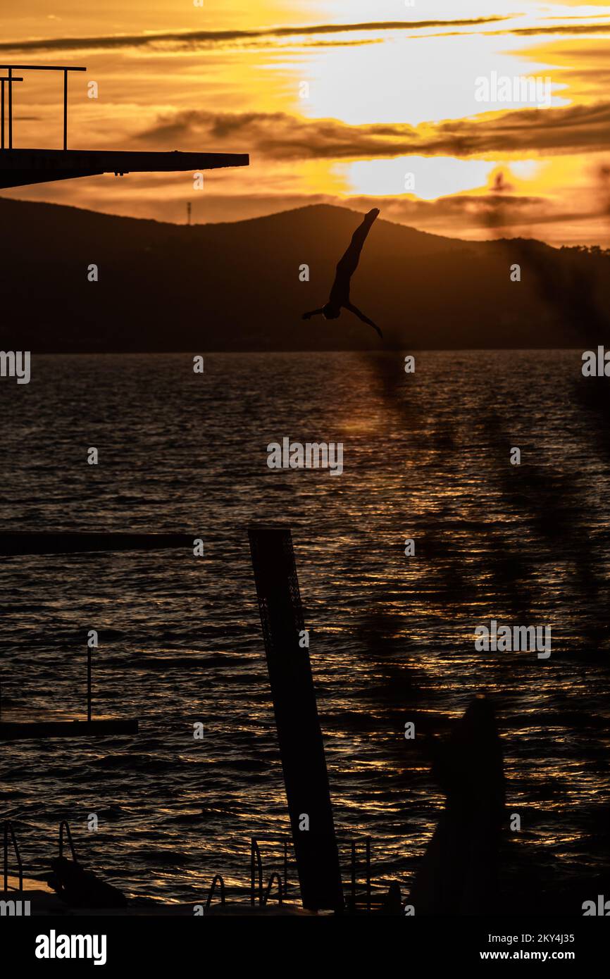 Boy jumps off the diving platform into the Adriatic Sea during sunset ...