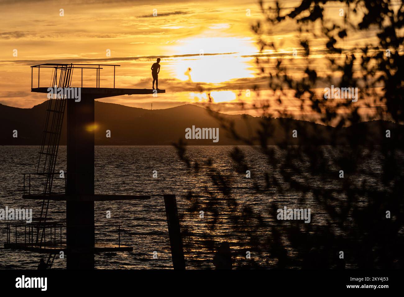 Boy jumps off the diving platform into the Adriatic Sea during sunset ...