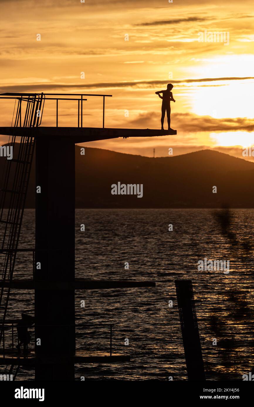 Boy jumps off the diving platform into the Adriatic Sea during sunset ...