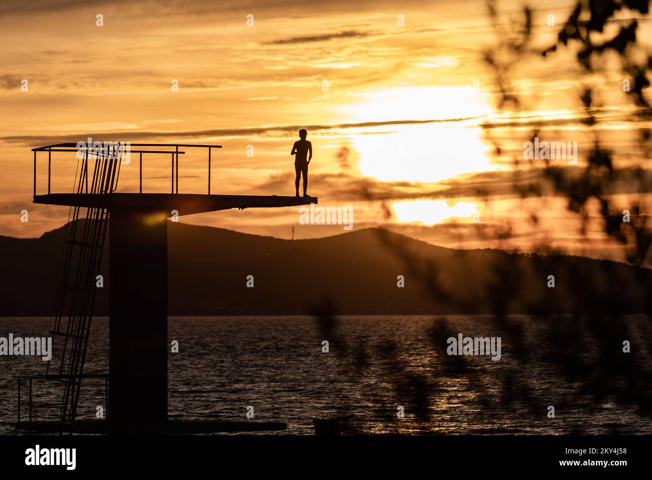 Boy jumps off the diving platform into the Adriatic Sea during sunset ...