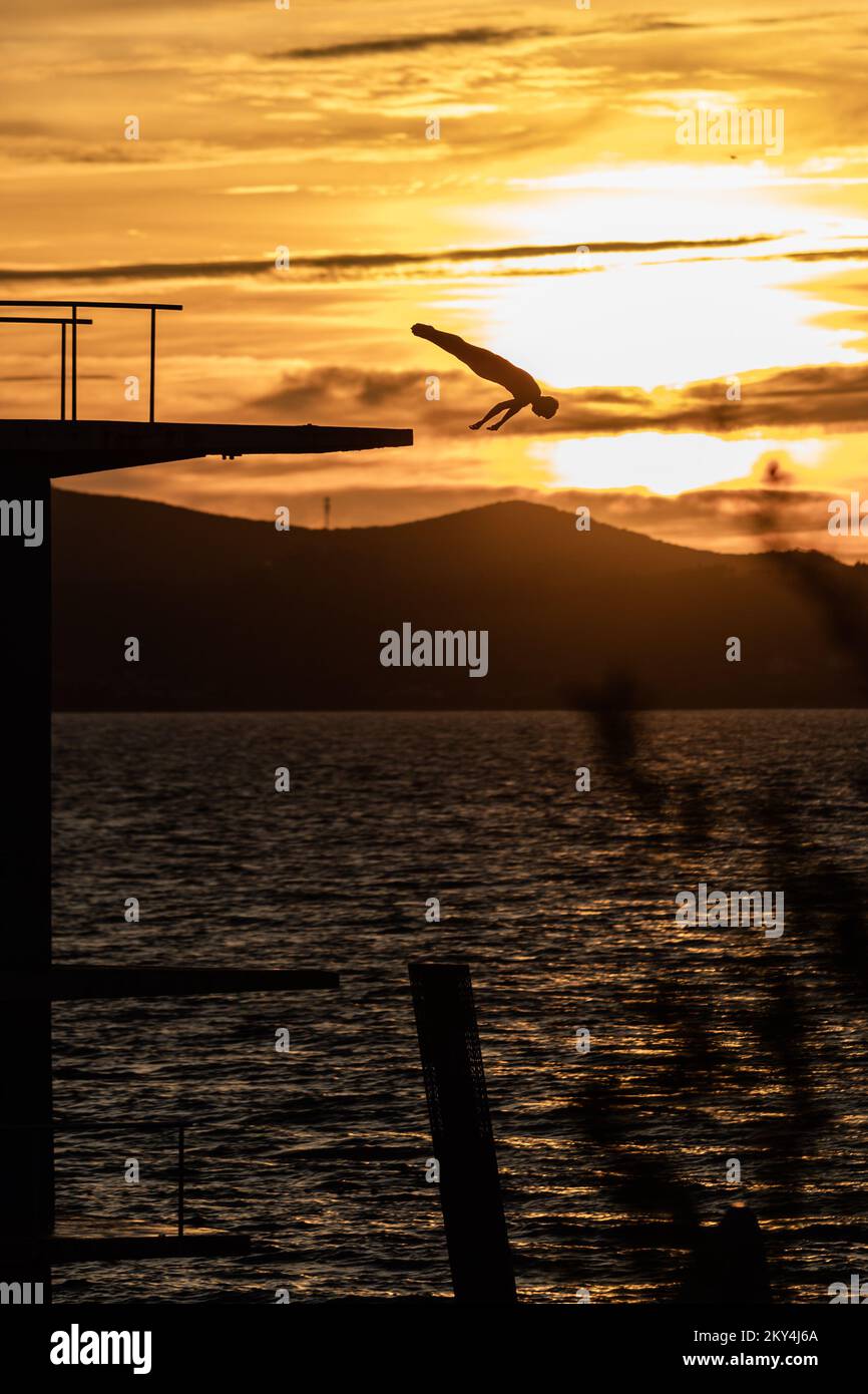 Boy jumps off the diving platform into the Adriatic Sea during sunset ...