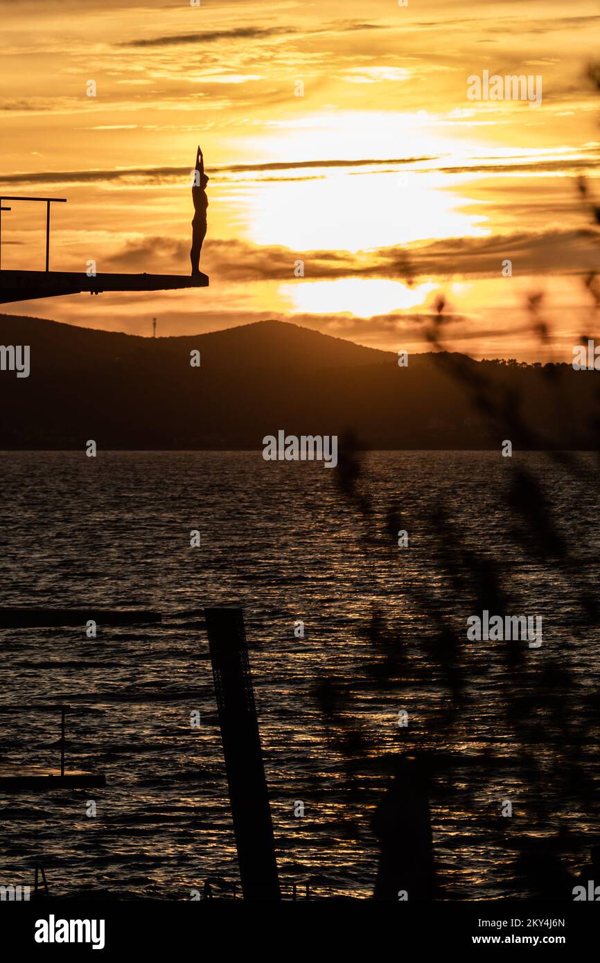 Boy jumps off the diving platform into the Adriatic Sea during sunset ...