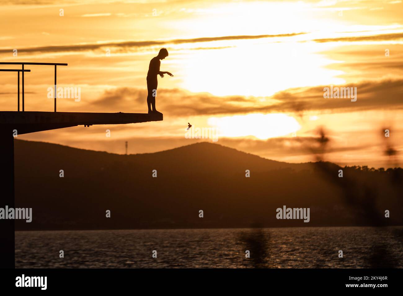 Boy jumps off the diving platform into the Adriatic Sea during sunset ...