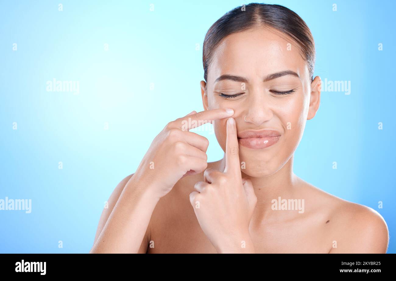 Woman, skincare and squeeze pimple on face with blue studio background ...