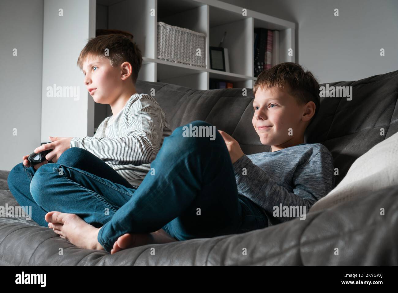 Cute school aged boys sit on sofa with game joysticks, from below view ...