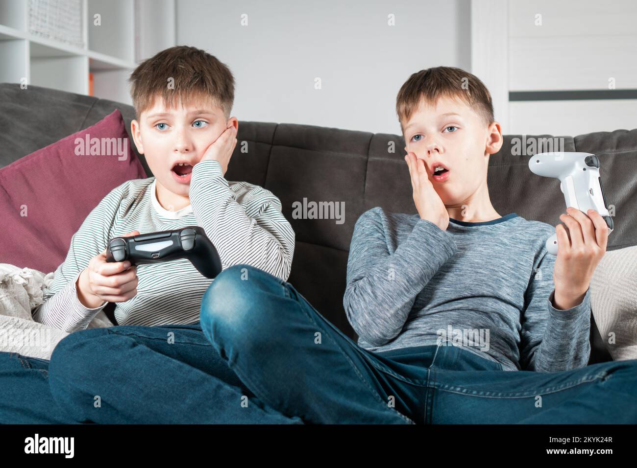 Shocked school aged boys sit on sofa with game joysticks. Children play ...