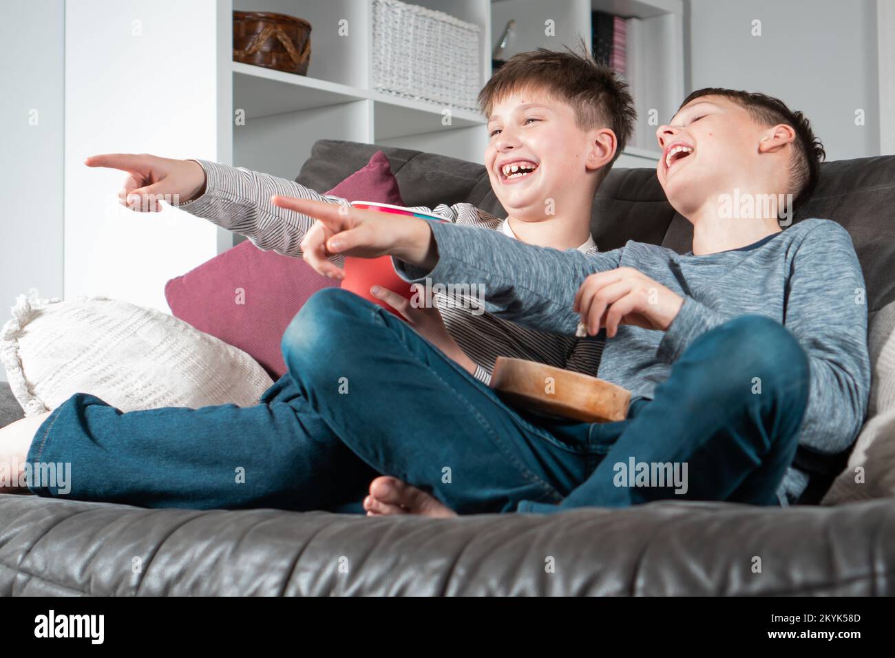 Cheerful school aged boys sit on sofa and laugh pointing finger forward ...