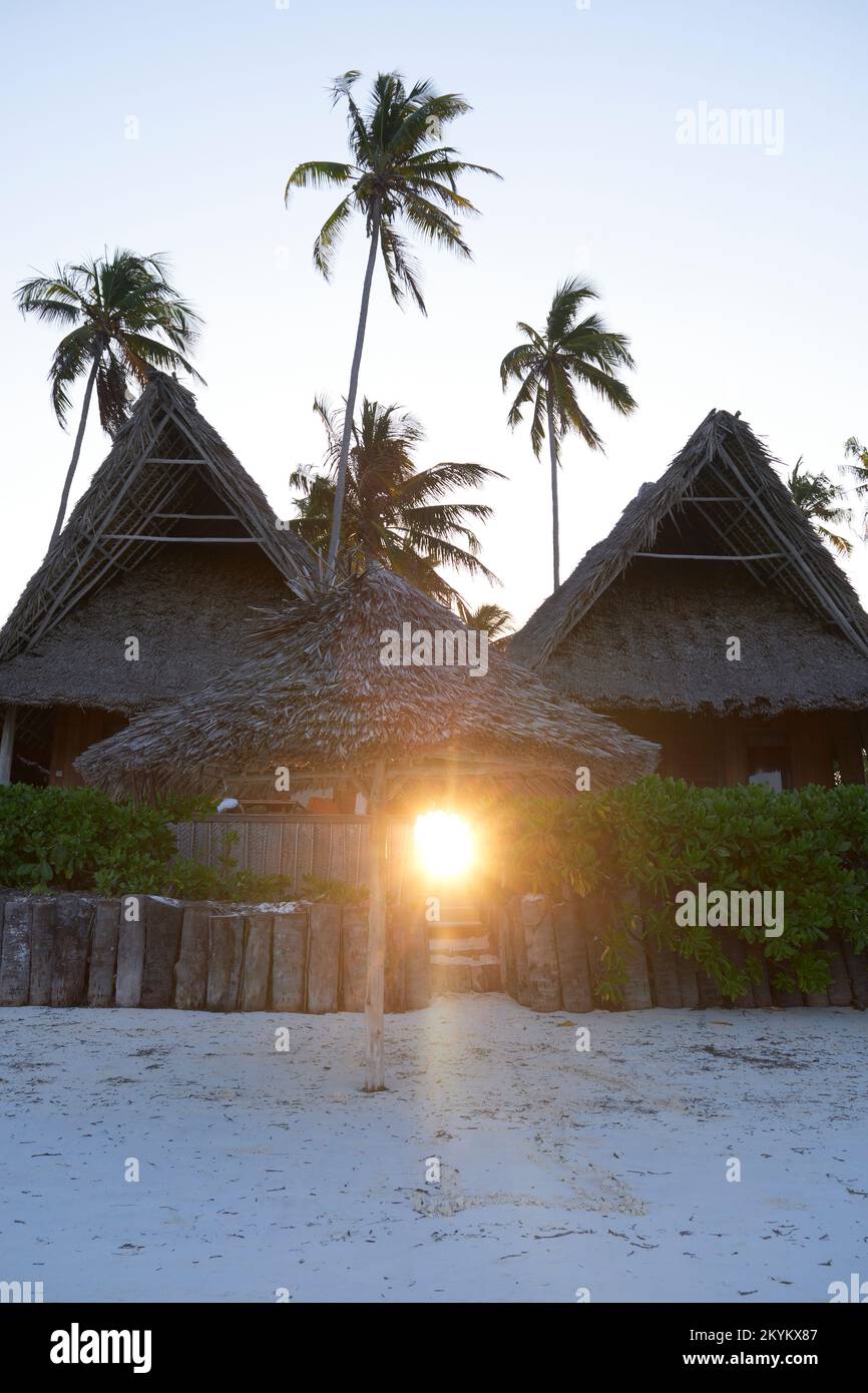 The sun sets behind palm trees and in between beach cottages during ...