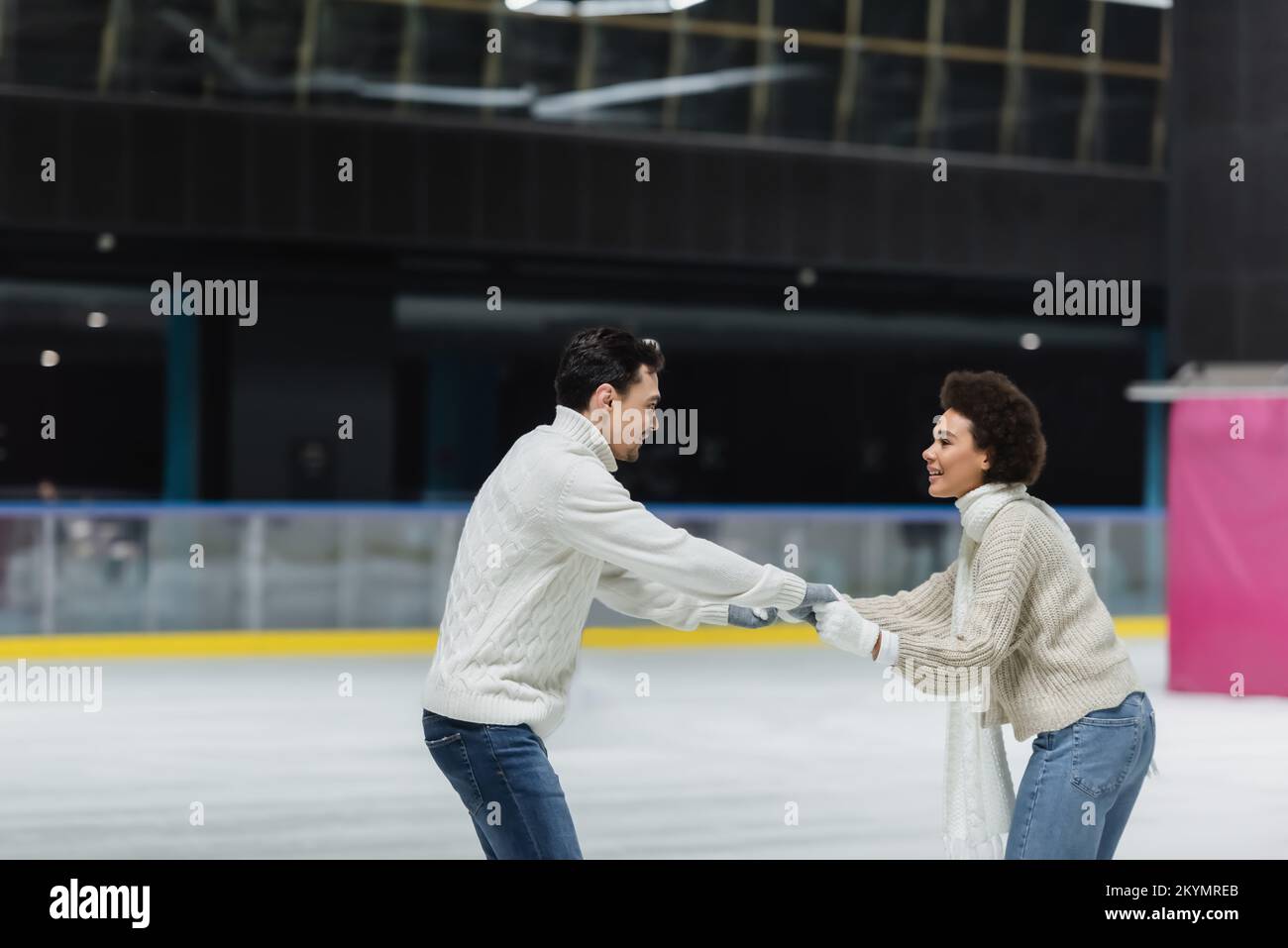 Side view of young interracial couple holding hands while ice skating ...