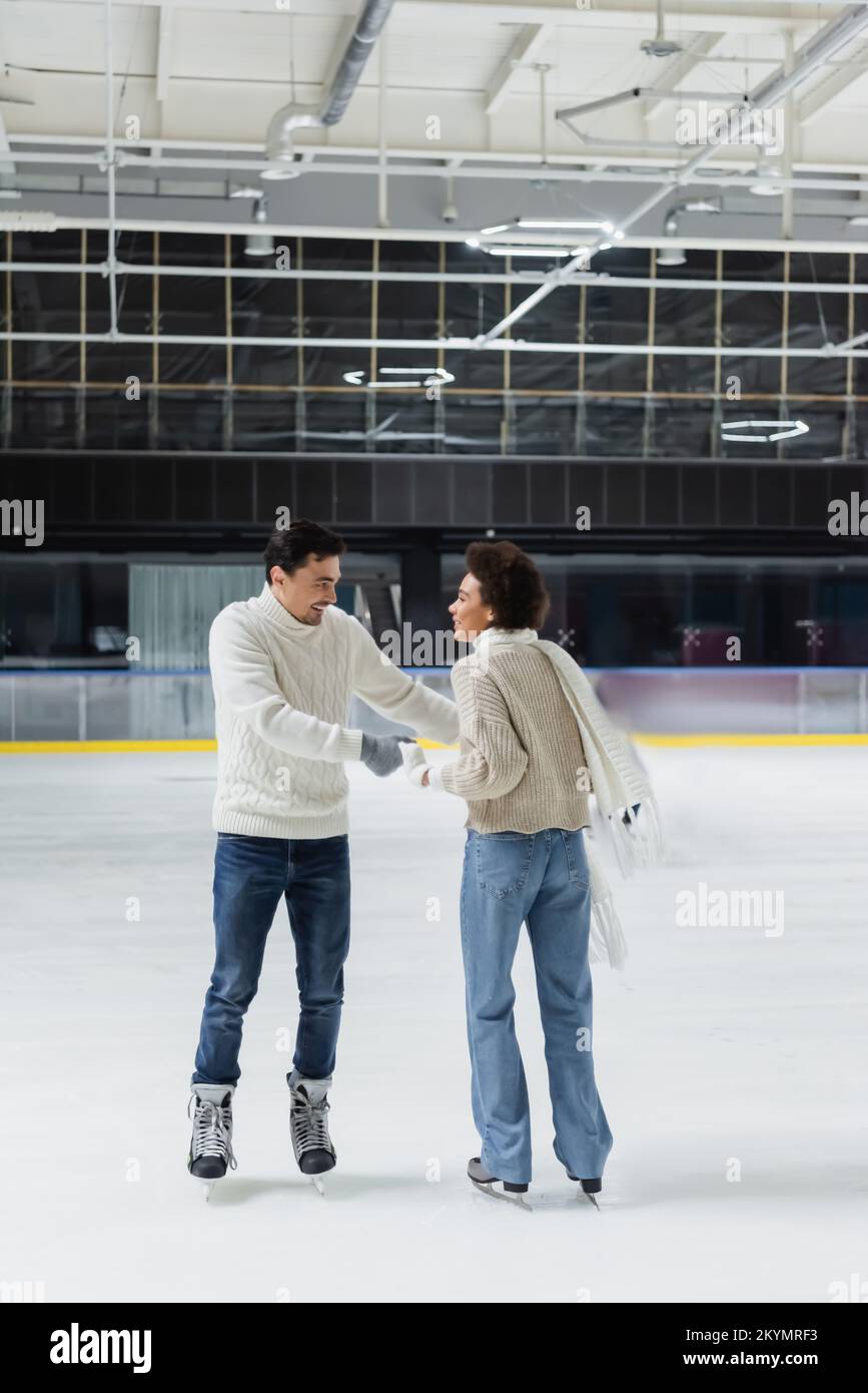 Positive multiethnic couple holding hands while skating on ice rink ...