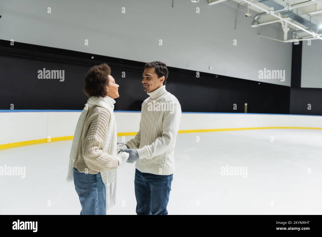 Multiethnic couple in warm knitted sweaters holding hands on ice rink ...