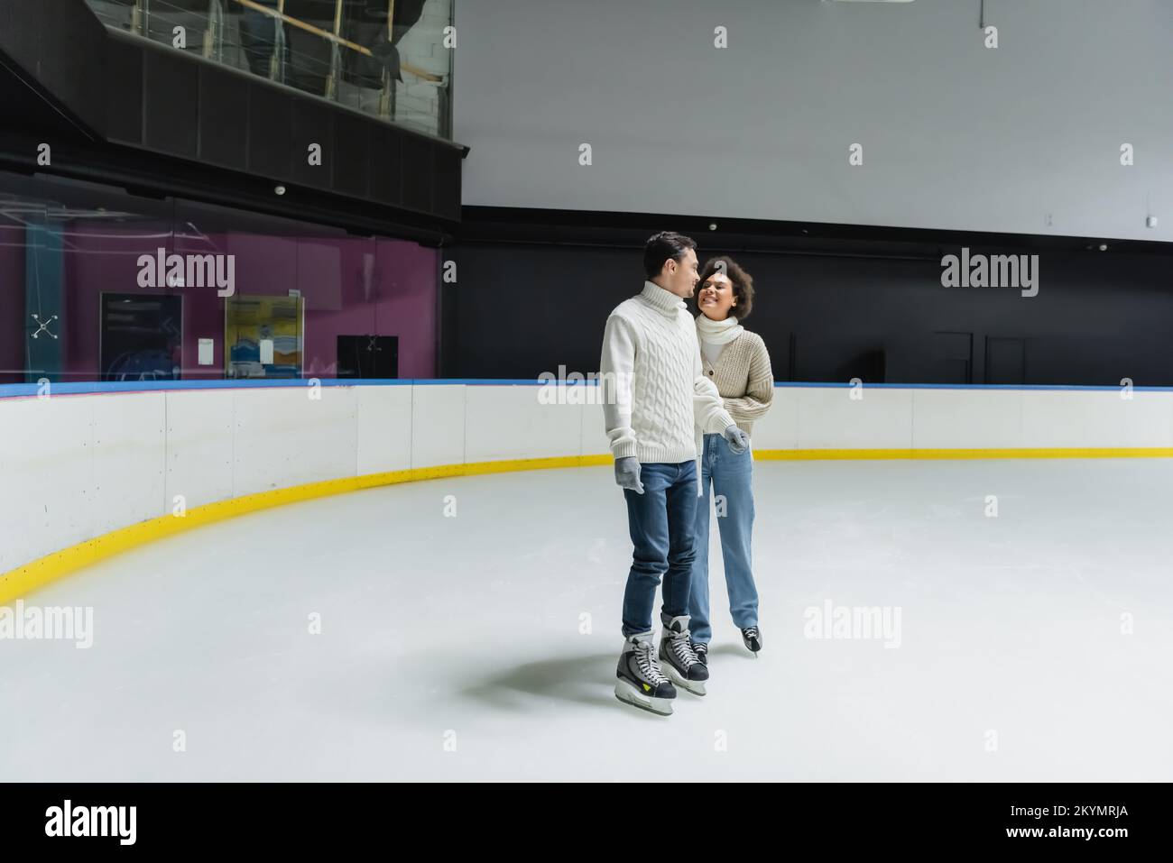Positive multiethnic couple in warm sweaters ice skating on rink Stock ...