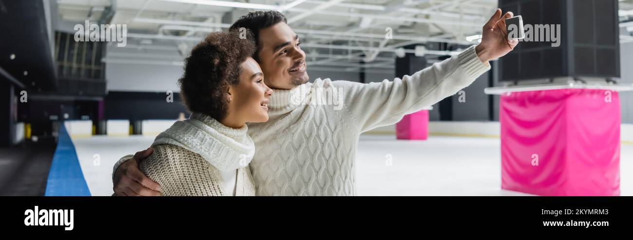 Positive interracial couple taking selfie on smartphone on ice rink ...