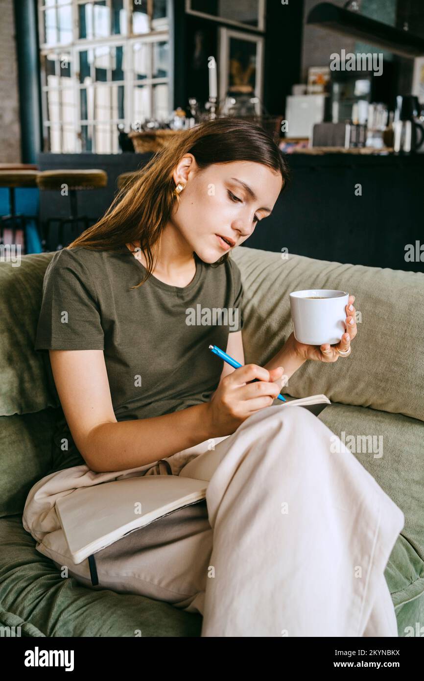 Young female entrepreneur writing in book holding coffee cup while ...
