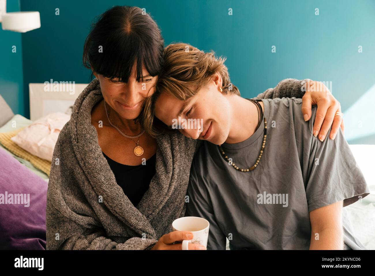 Smiling mother with arm around son leaning on shoulder sitting on bed ...