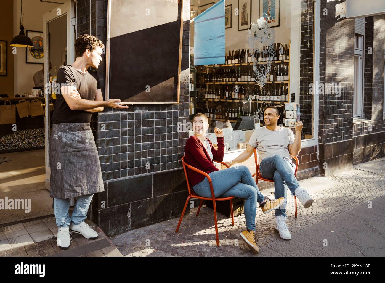 Male owner talking to customers sitting on chair at sidewalk outside ...