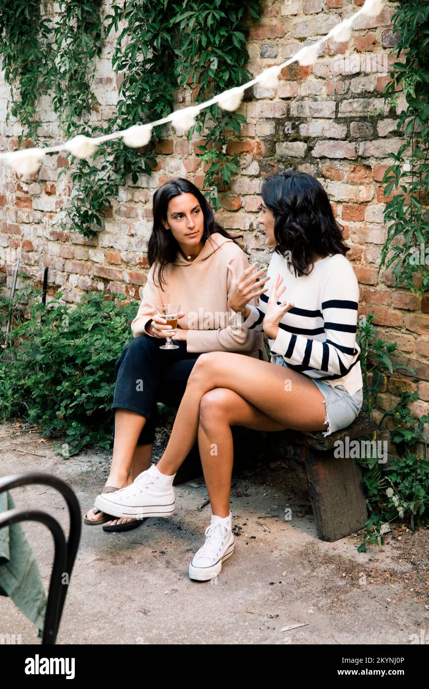 Female friends talking to each other while sitting on bench near brick ...