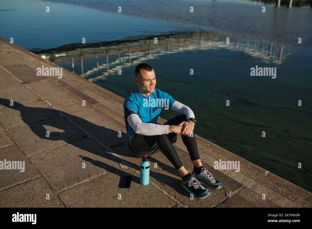 Athletic man runner sitting rests after training on city river bank ...