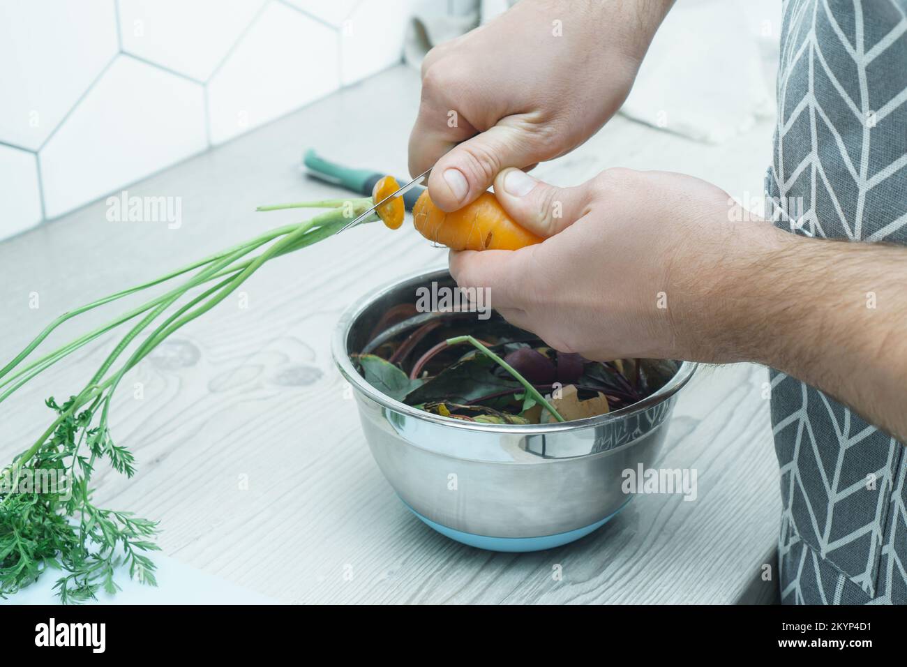 Male hands peeling fresh carrot with knife and cutting haulm over bowl of vegetable peelings ...