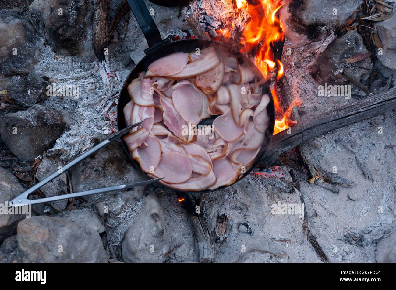 Australian style bacon being cooked over a camfire with silver tongs ...