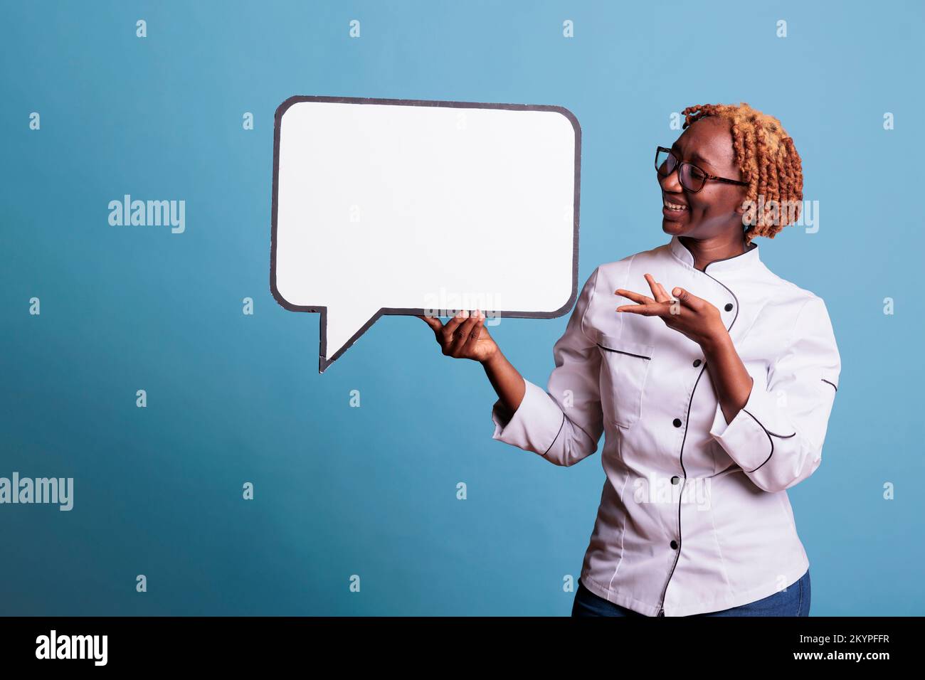 Woman smiling kitchen staff member pointing to blank dialogue cloud with copy space. Cook ...