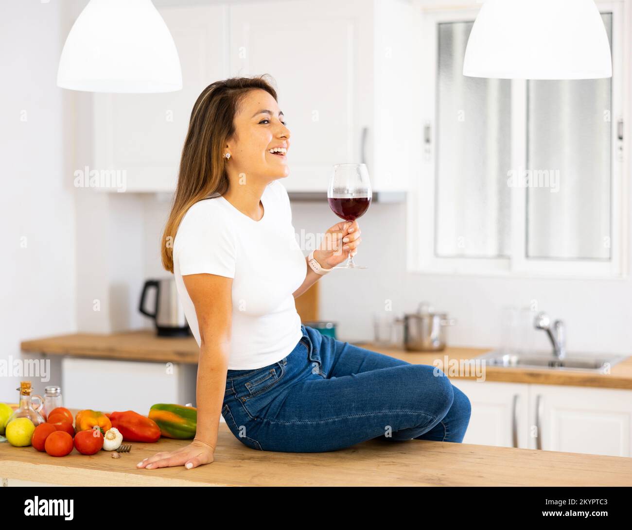 Smiling young Hispanic woman drinking wine sitting on table in kitchen ...