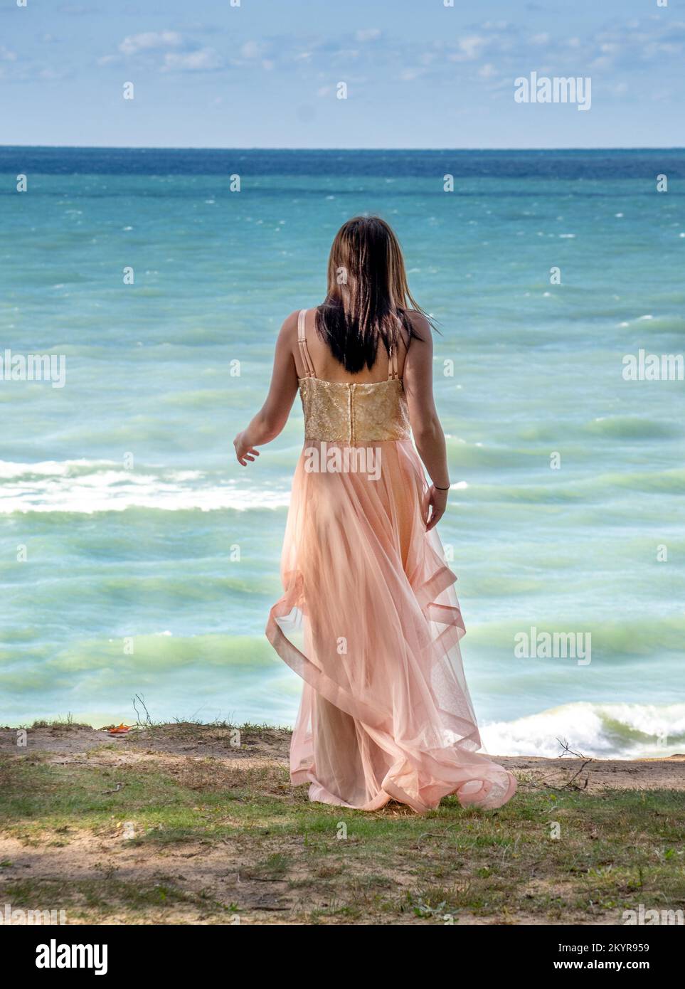 A young woman in a flowing formal dress, stands on a ridge high above ...