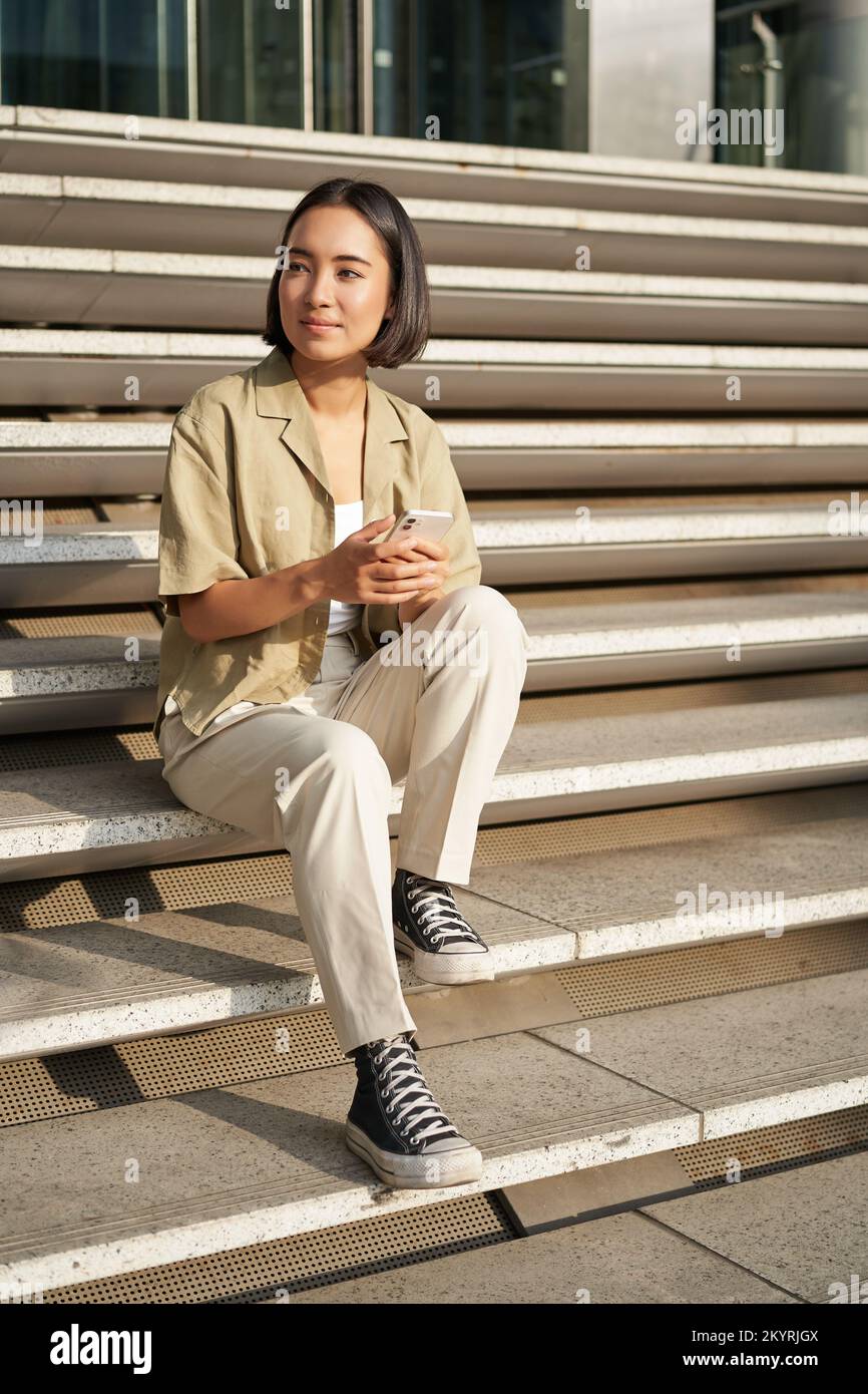 Beautiful asian girl sitting on stairs outside building, using mobile ...