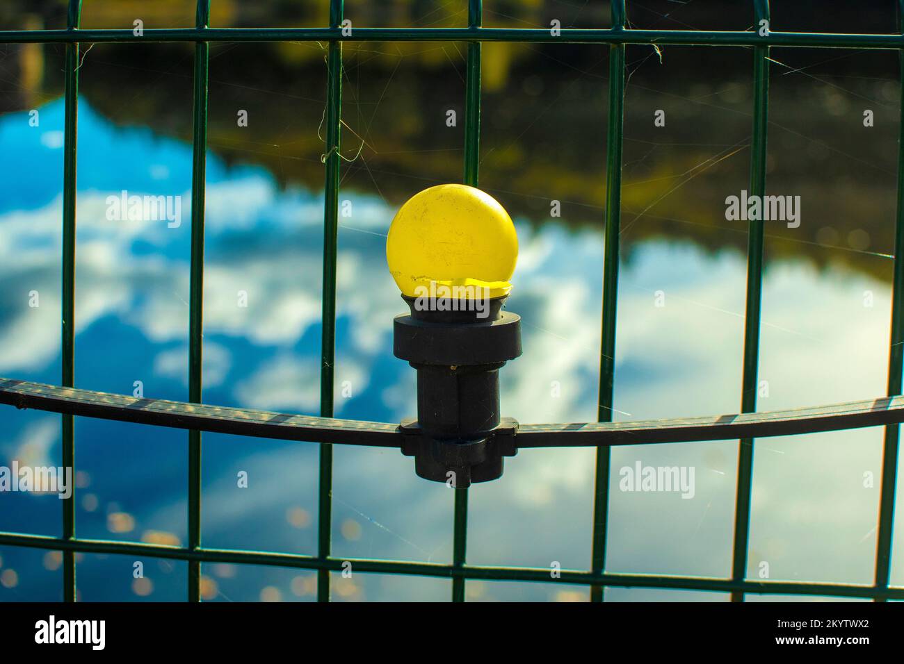 Yellow light bulb hanging near a fence reflecting sky and water in a ...