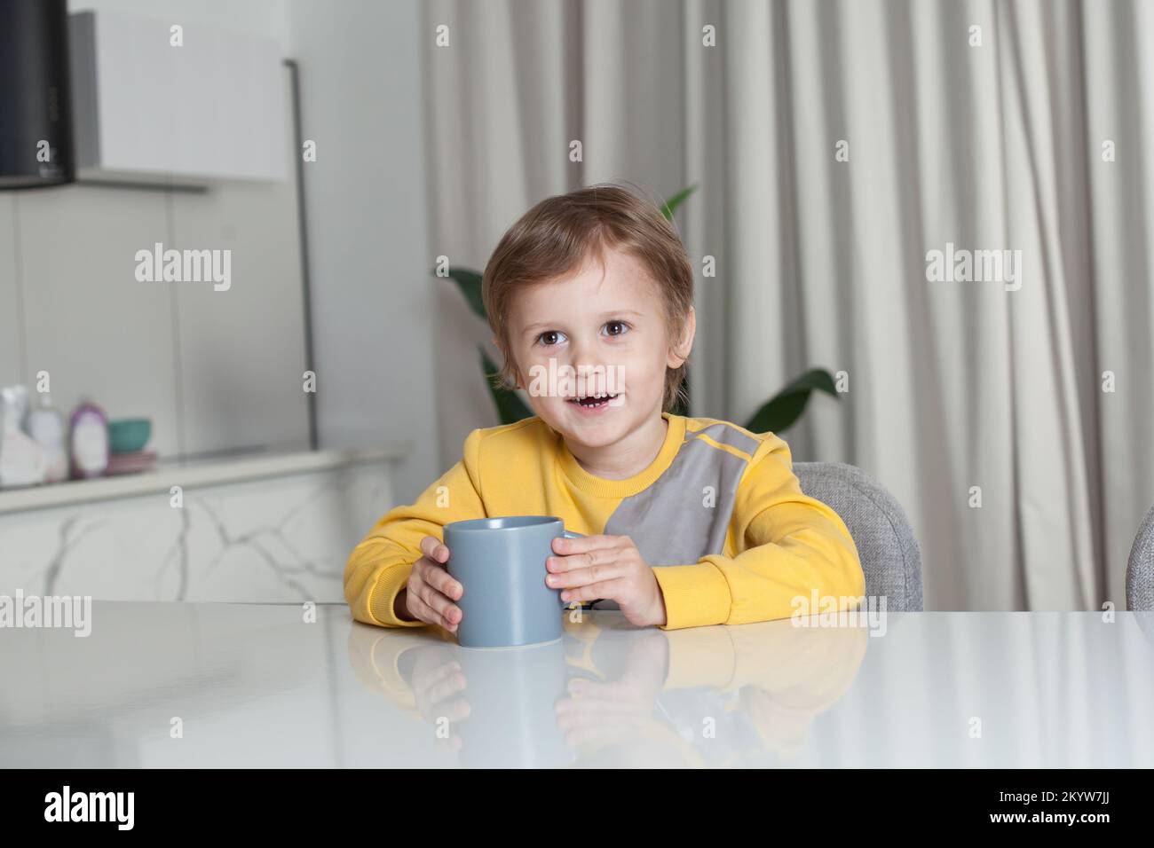 Happy cute child boy in yellow sweater sitting by the table and holding ...