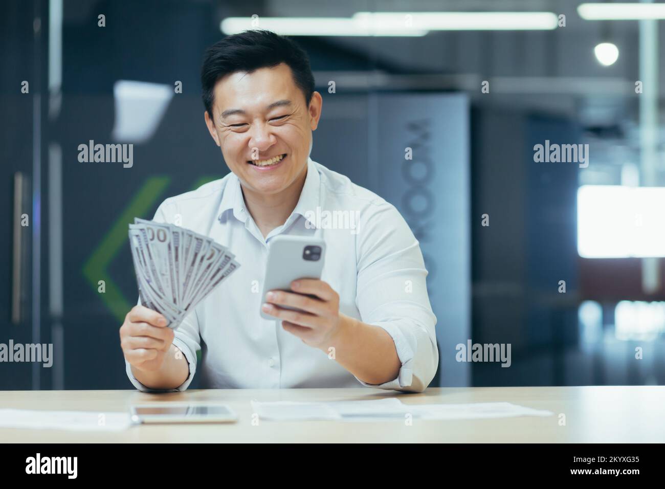 Happy young Asian man sitting in the office at the table holding cash ...