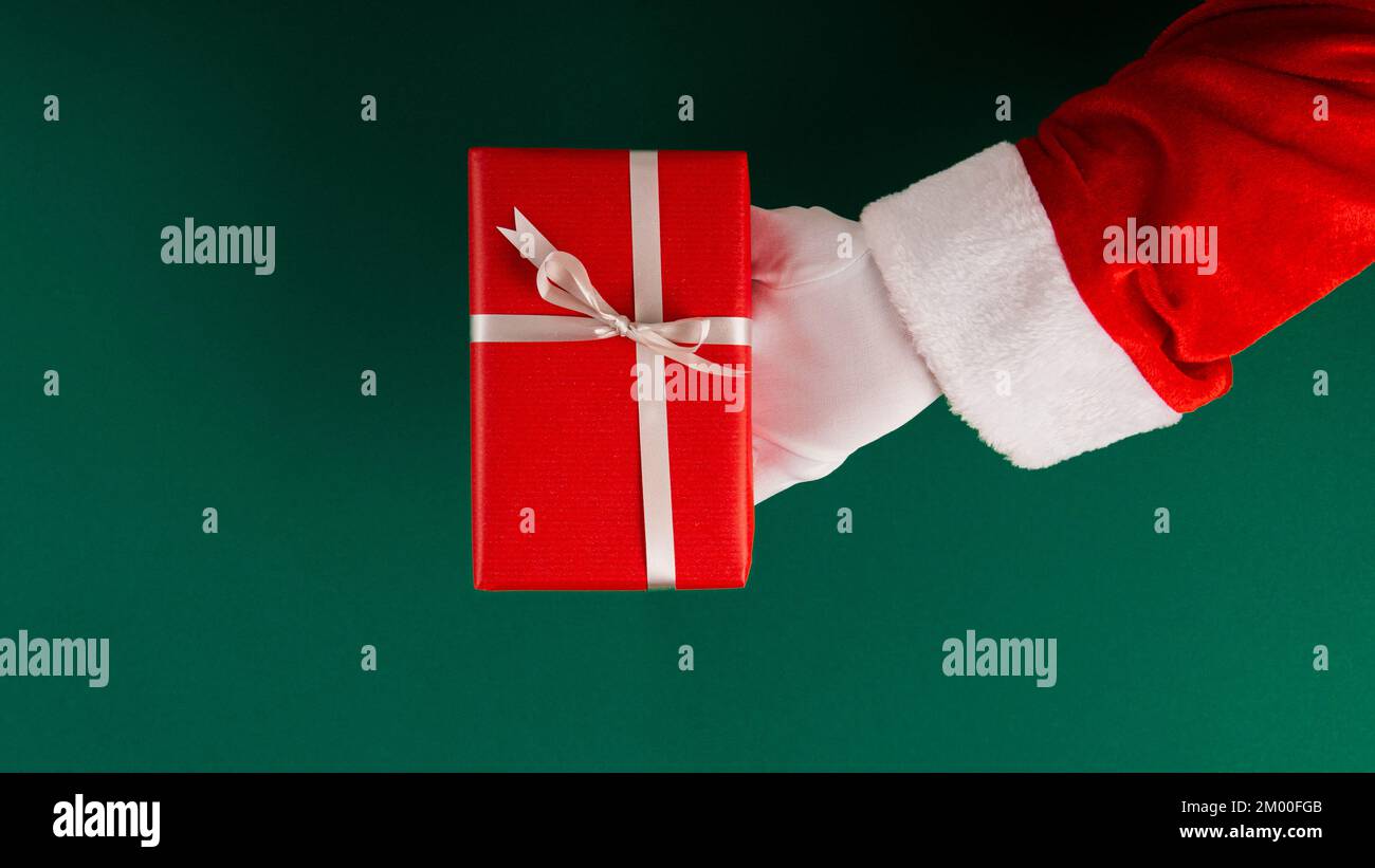 Hand of santa claus gives a red wrapped box with ribbon on a green ...