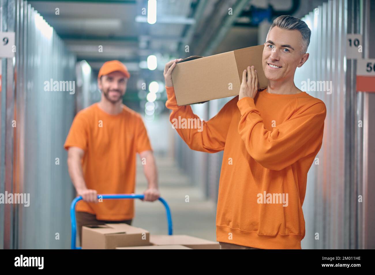 Cheerful middle-aged Caucasian loader and his male colleague unloading ...