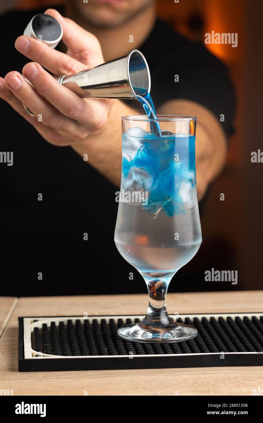 Professional male bartender pouring a blue alcoholic drink from the jigger on cocktail glass ...