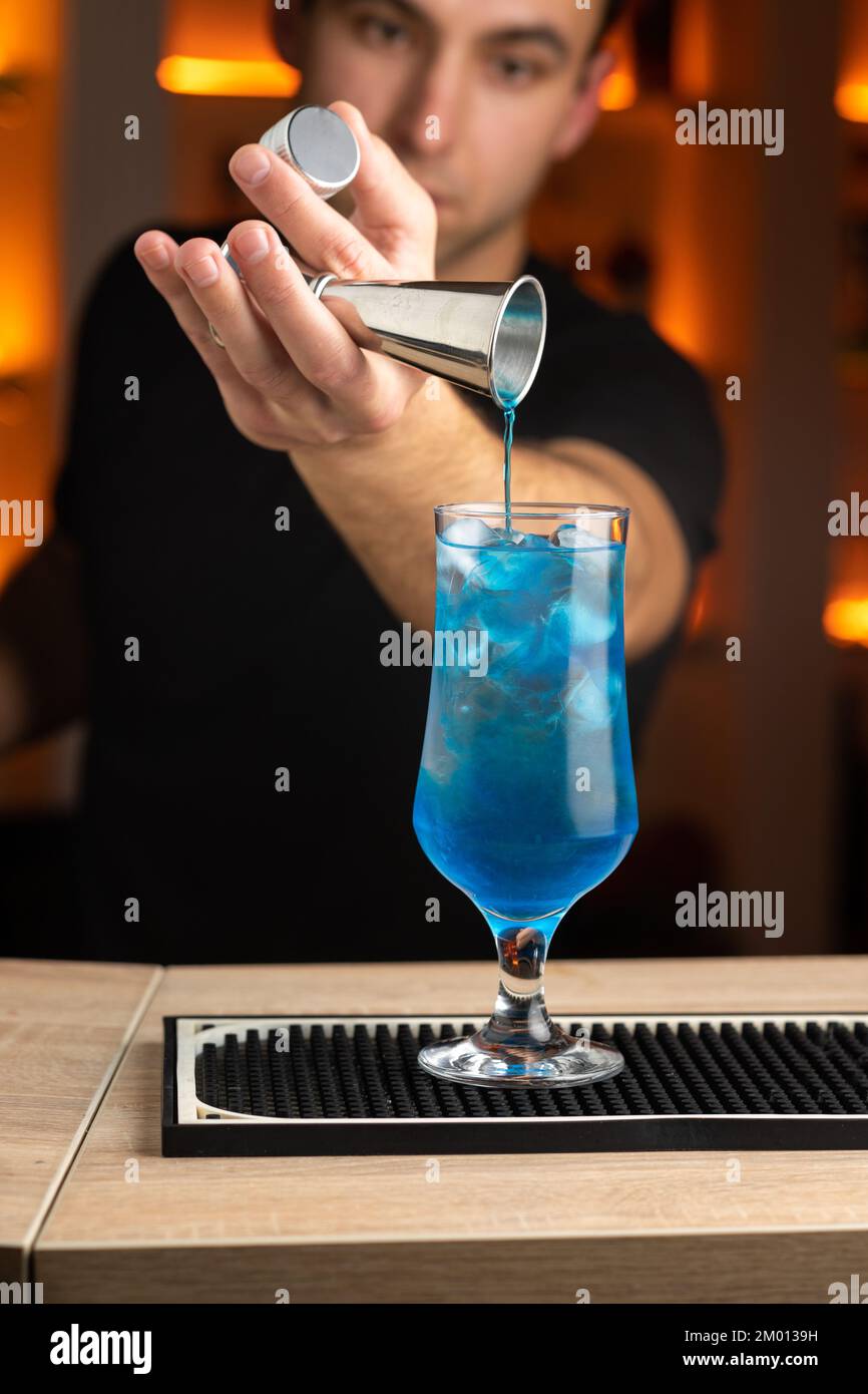 Professional male bartender pouring a blue alcoholic drink from the jigger on cocktail glass ...