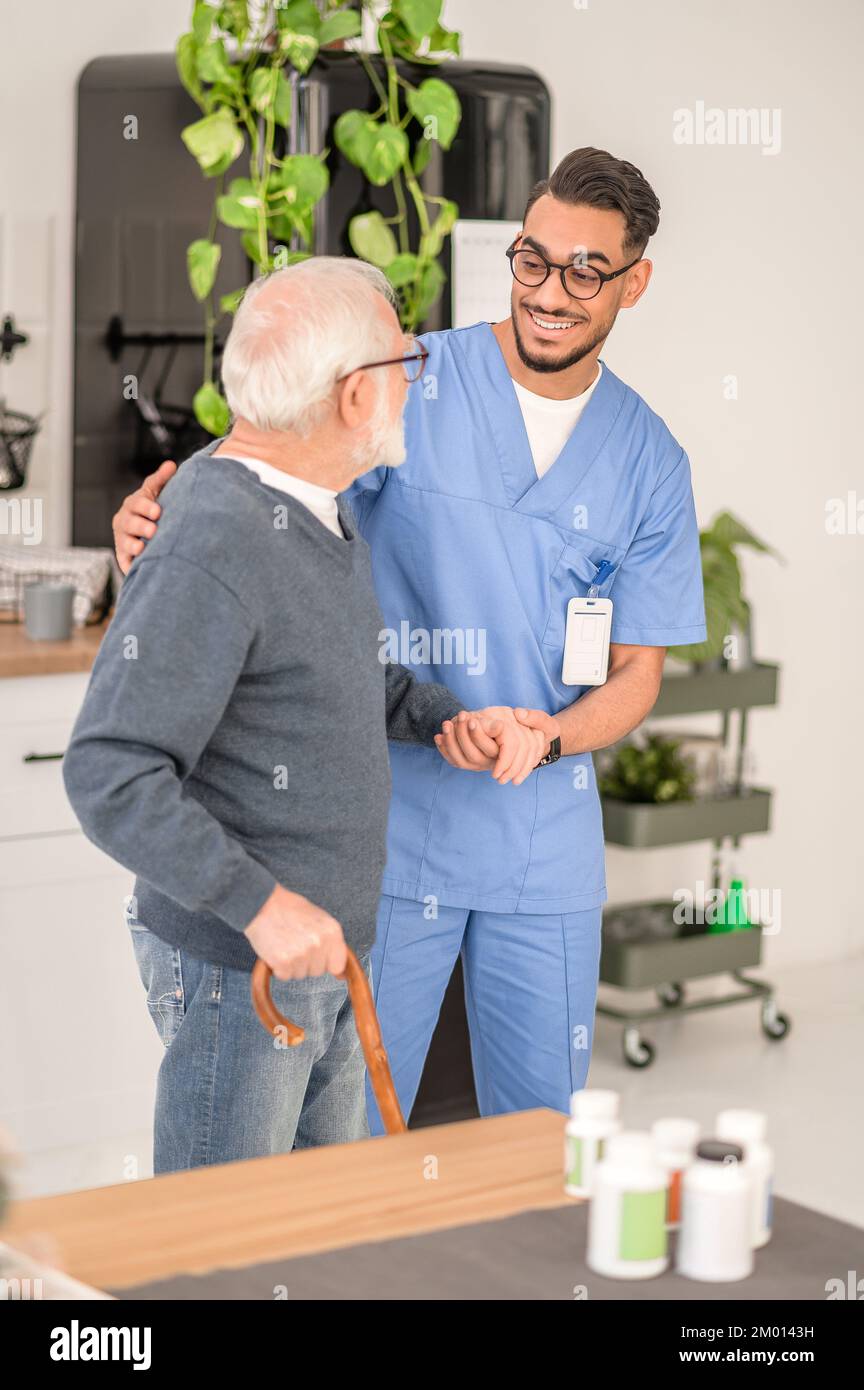 Smiling amiable young in-home nurse assisting his male patient to lean ...