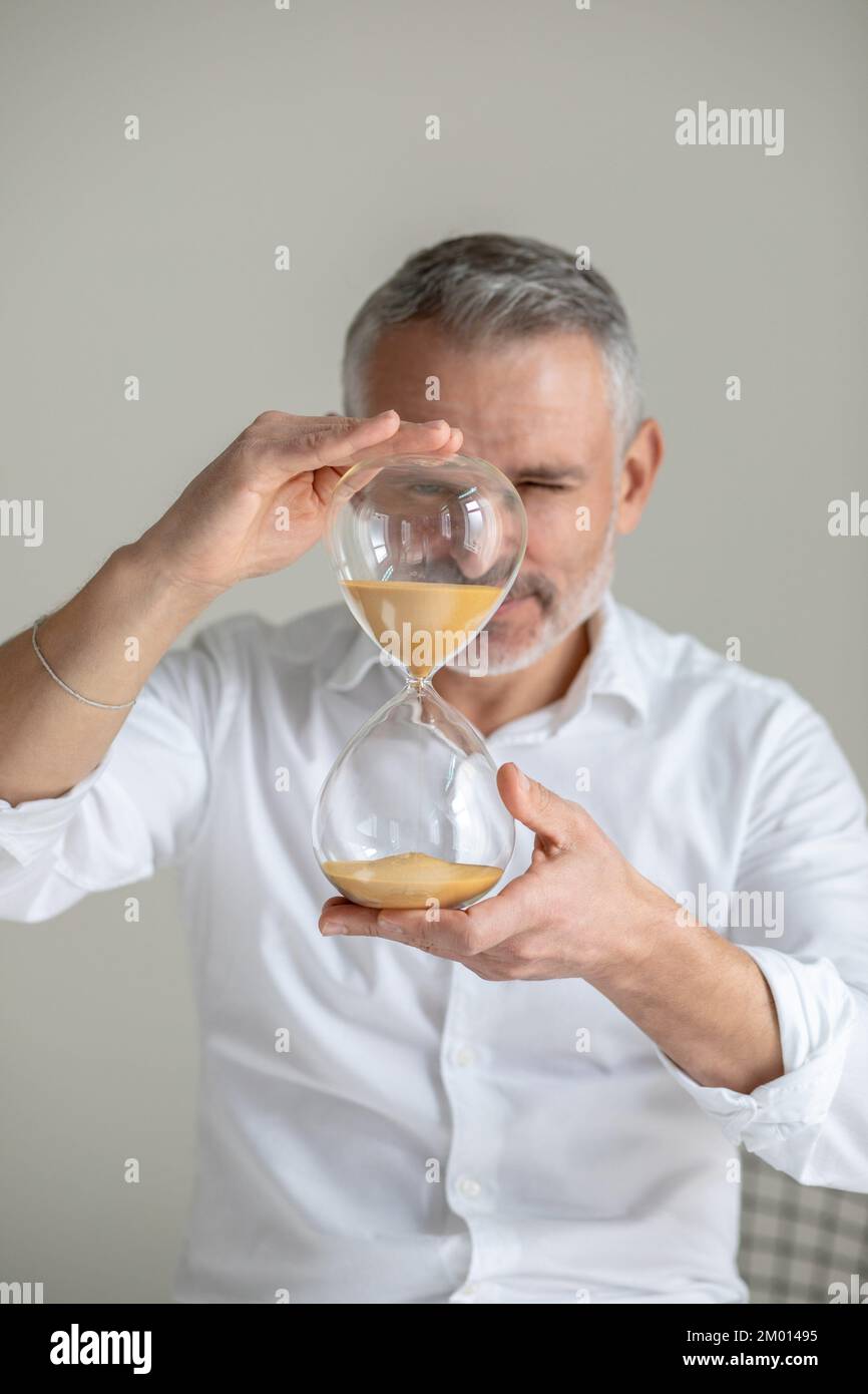 Time. Gray-haired smiling man squinting his eye holding sand watch at ...